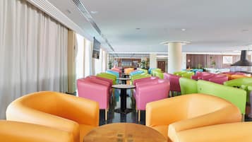 Um saguão de hotel com assentos coloridos, uma mesa central e vista para a área de refeições.