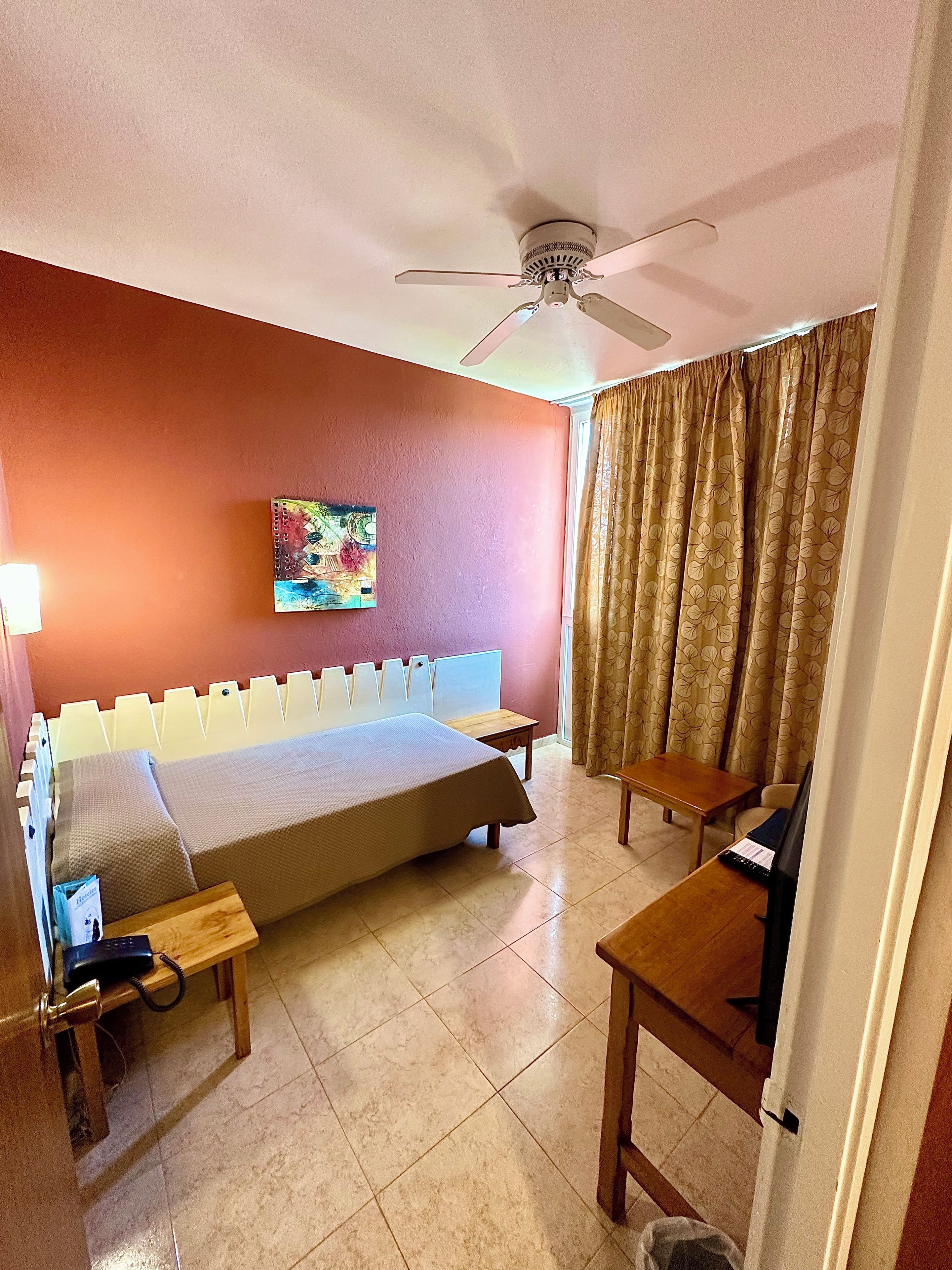 Quarto de hotel com cama, escrivaninha, cadeira, ventoinha no teto, quadro na parede e janela com cortinas.