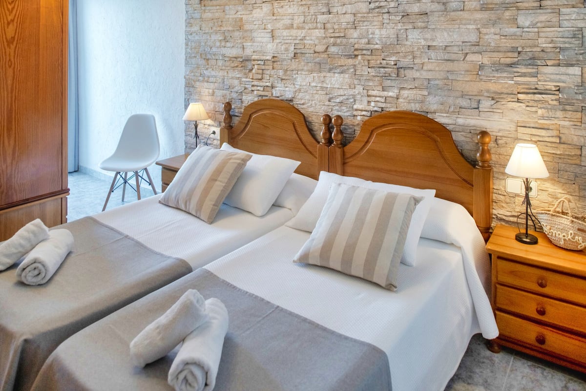 Quarto de hotel com duas camas, parede de pedra, mesa de cabeceira de madeira, abajur e uma cadeira.