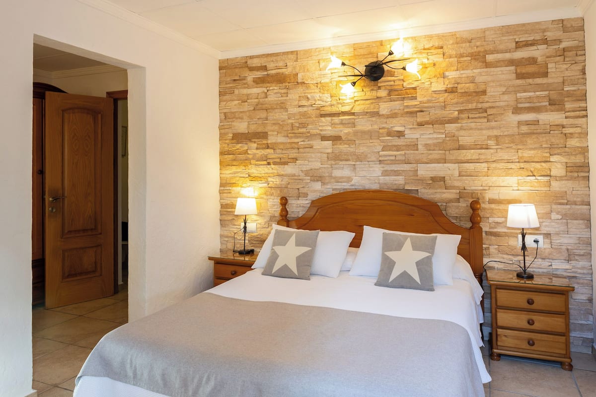 Uma cama bem arrumada com cabeceira de madeira, duas mesas de cabeceira com luminárias e parede com textura de pedra atrás da cama.