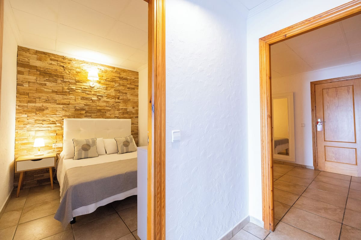 Quarto com parede de pedra, uma cama com cabeceira, mesa de cabeceira com abajur e porta de madeira.