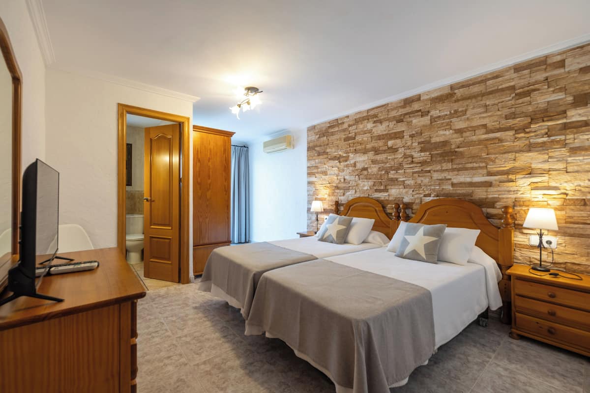 Quarto com duas camas, uma mesa de cabeceira de madeira, uma escrivaninha com computador e uma parede de pedra.