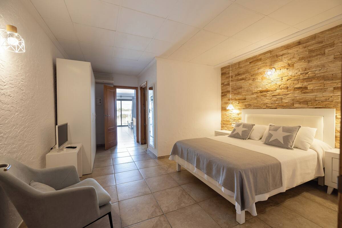 Quarto moderno com uma cama grande, uma mesinha de cabeceira, uma cadeira e uma televisão sobre um pequeno móvel.