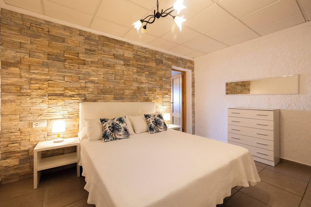 Quarto com parede de pedra, cama com roupa de cama branca, mesa de cabeceira com abajur e cômoda com gavetas.