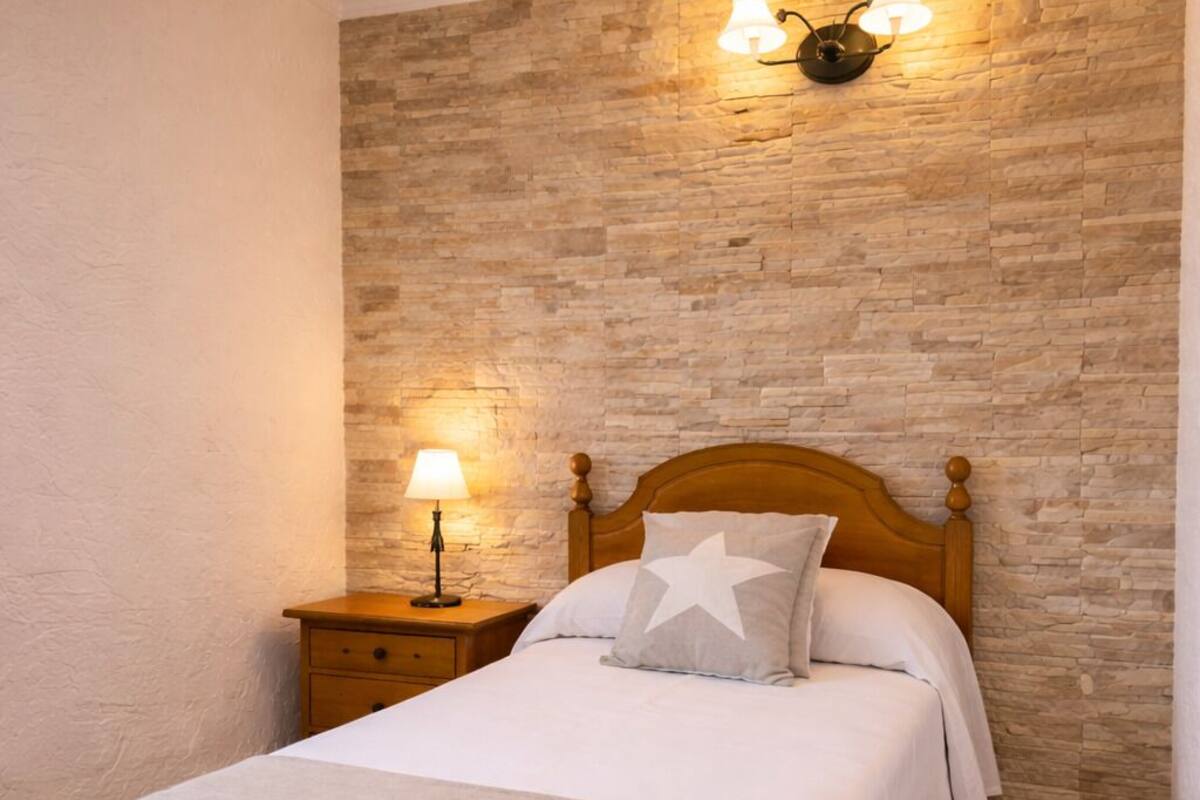 Quarto com parede de pedra, estrutura de cama de madeira, mesa de cabeceira com abajur e travesseiro em formato de estrela.