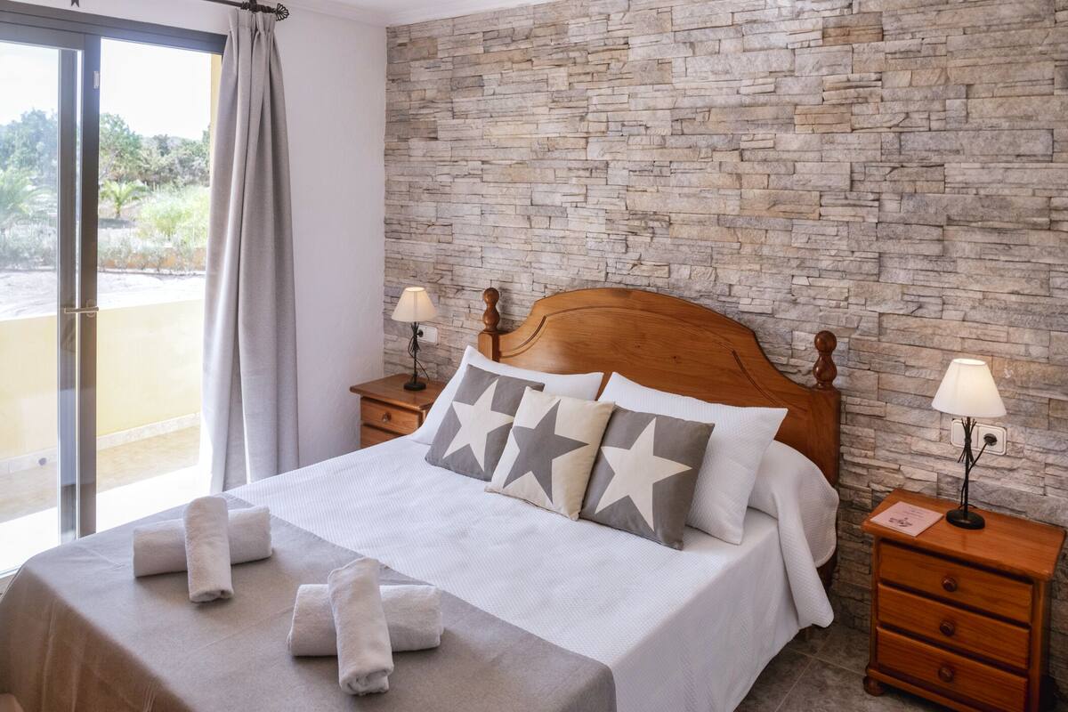Uma cama bem arrumada com almofadas estampadas em formato de estrela, cabeceira de madeira e parede de pedra ao fundo.