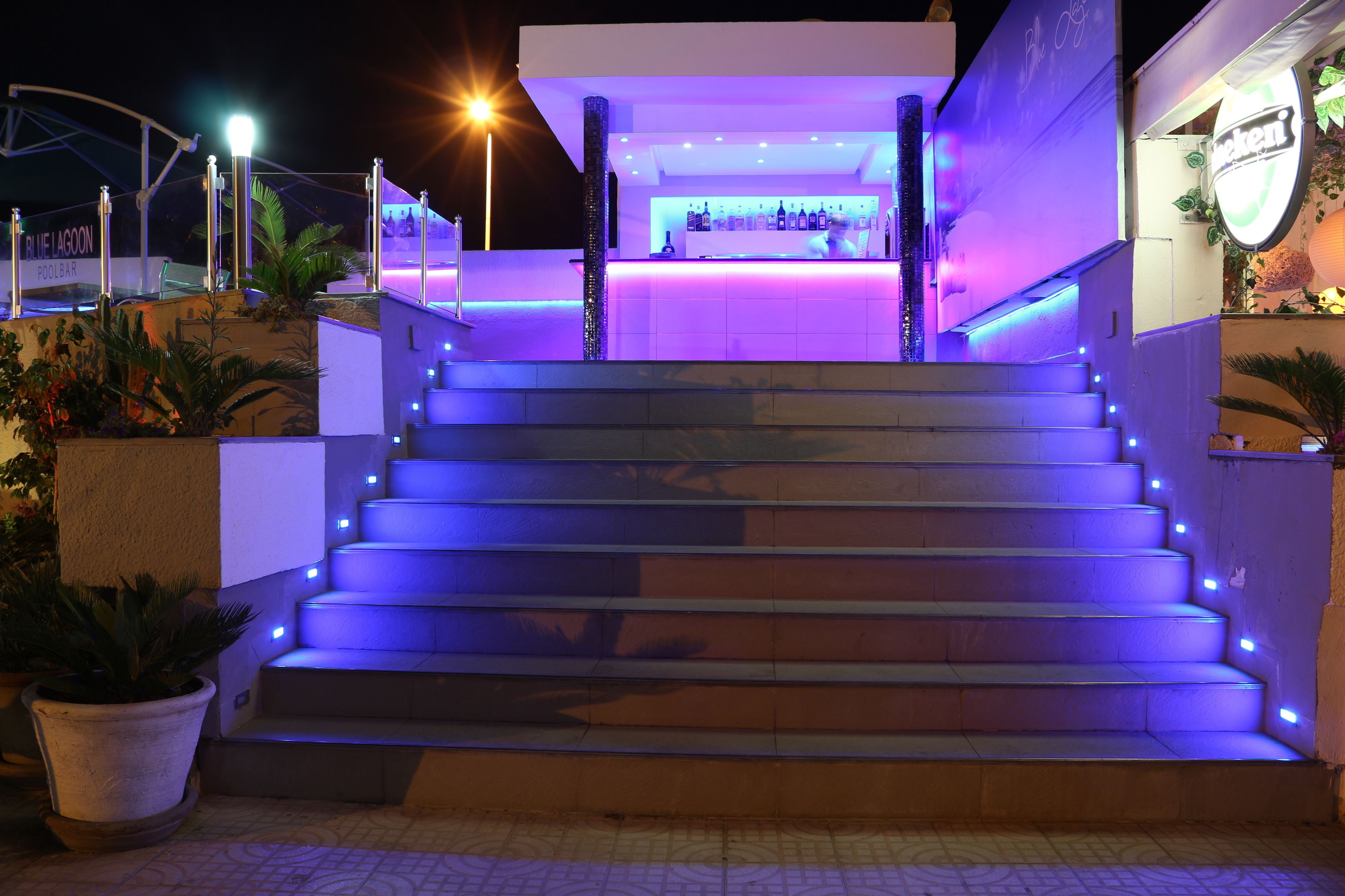 Escadaria iluminada com luzes LED azuis que leva a uma área de bar, com um aviso de piscina ao fundo.