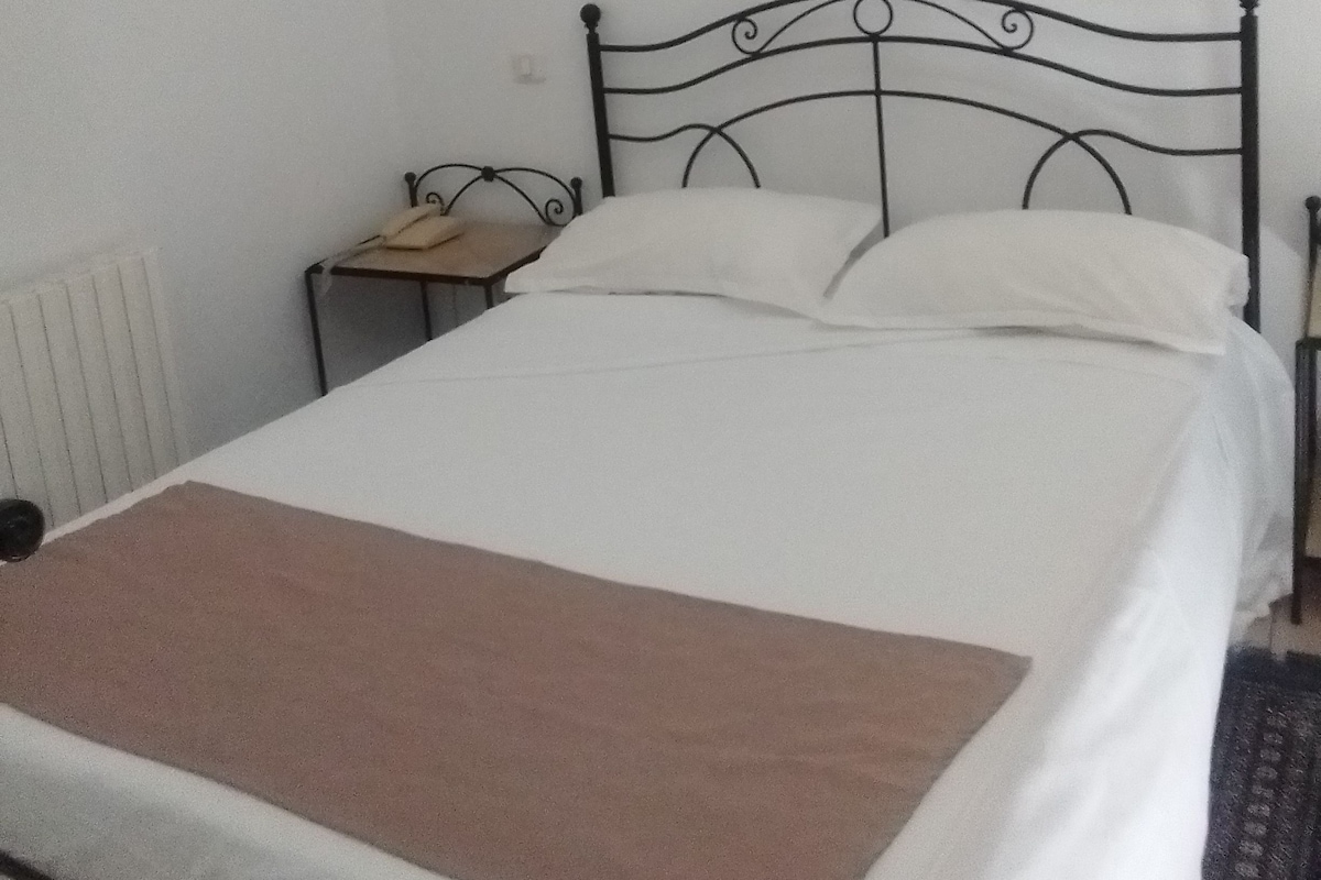 Uma cama de solteiro com estrutura de metal, uma mesa de cabeceira com abajur e um quadro na parede.