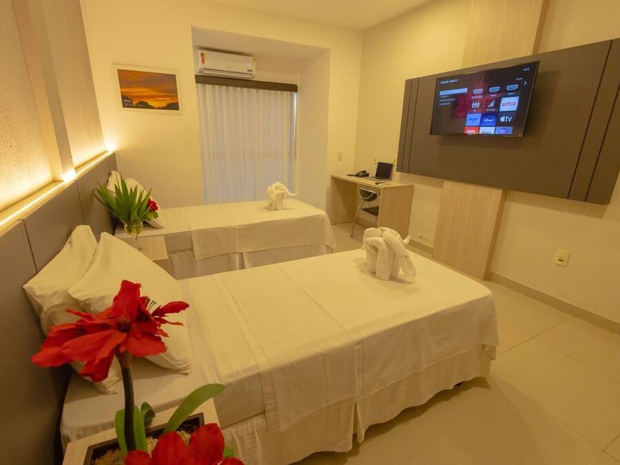 Quarto de hotel com duas camas, uma TV fixada na parede e uma pequena escrivaninha com uma cadeira.