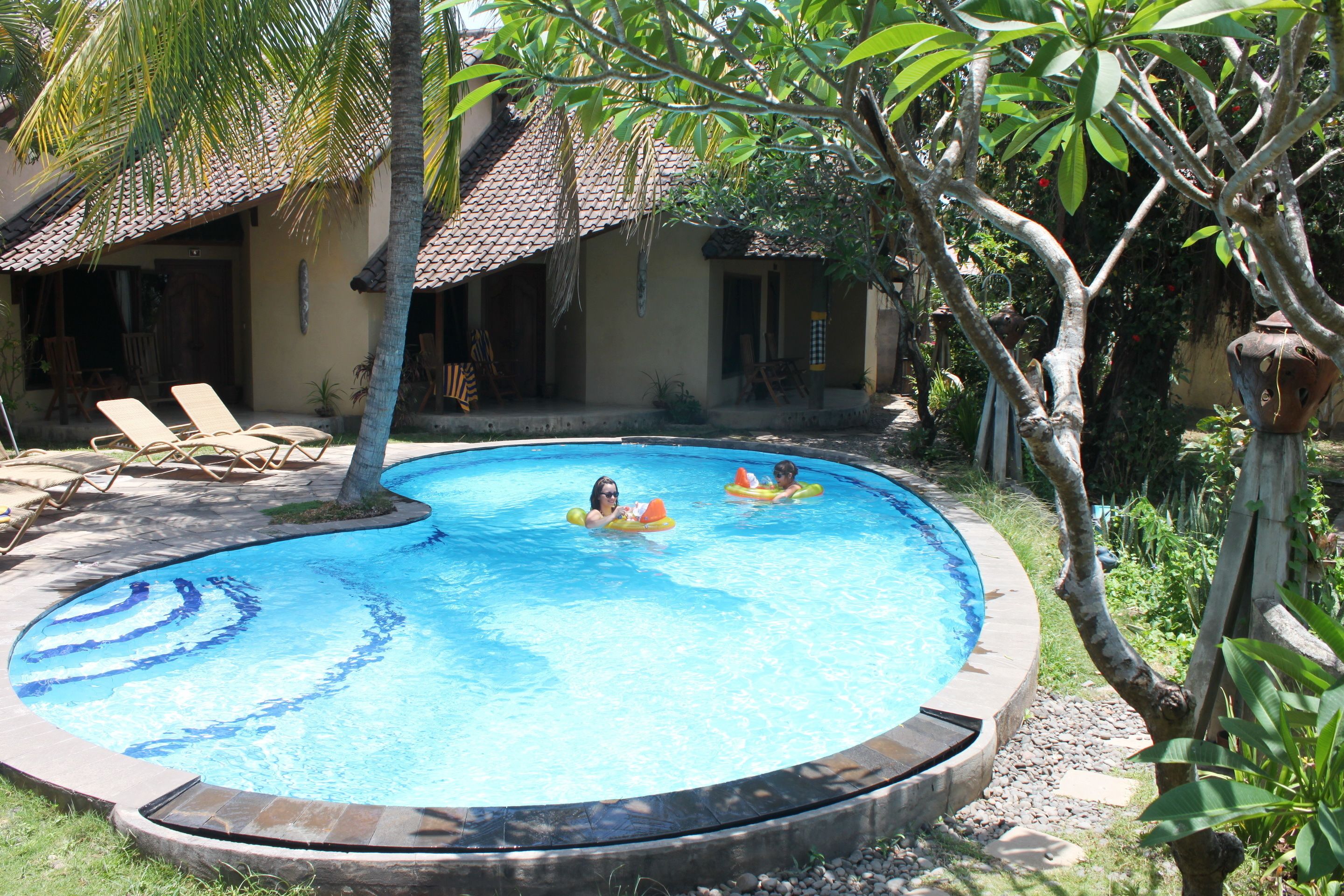 Piscina com duas pessoas em boias, cercada por palmeiras e uma casa com telhado de azulejos.