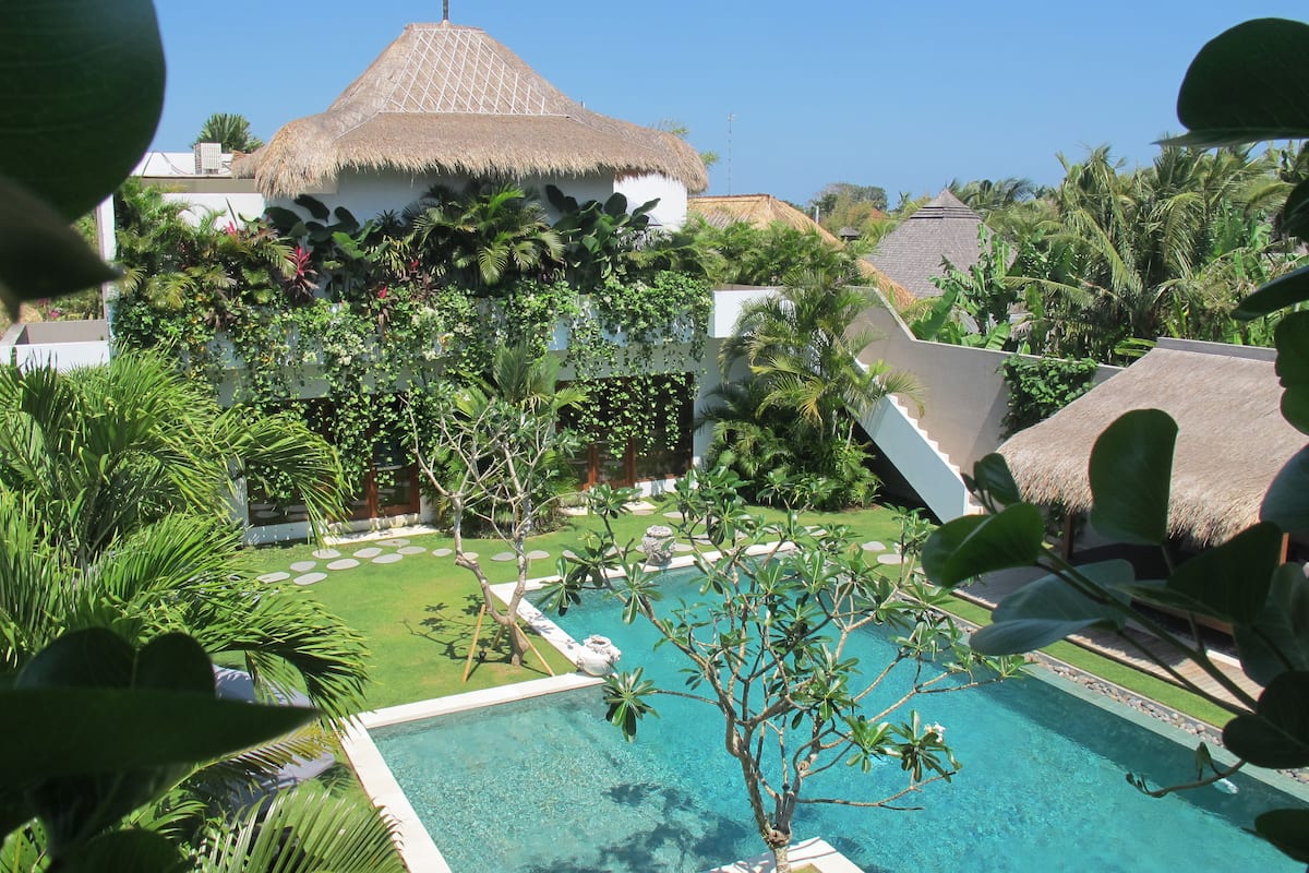 Piscina cercada por vegetação exuberante, um edifício com telhado de palha e uma parede branca coberta por plantas trepadeiras.