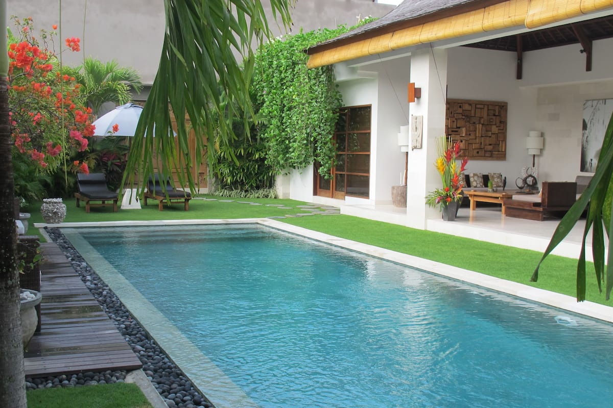 Piscina com deck de madeira, cercada por gramado bem cuidado e plantas tropicais.