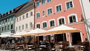 Uma fileira de mesas de café ao ar livre com guarda-sóis, um prédio vermelho com a placa 'Hotel' e um prédio verde com a placa 'Engbers'.