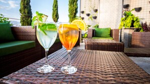 Duas bebidas sobre uma mesa de vime ao ar livre, com assentos verdes e árvores altas ao fundo.