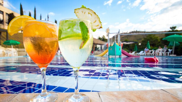 Dois cocktails sobre uma mesa de azulejos à beira da piscina, com piscina e guarda-sóis ao fundo.