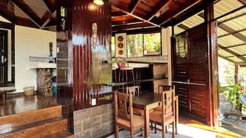 Um quarto espaçoso com teto alto, vigas de madeira, área de refeições com cadeiras e mesa, e vista para o exterior através de grandes janelas.