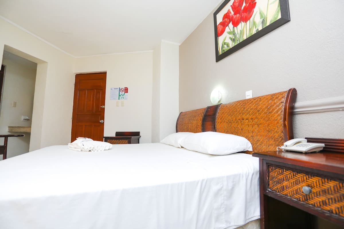 Quarto de hotel com uma cama, uma mesa de cabeceira com abajur, cabeceira de madeira e um quadro na parede.