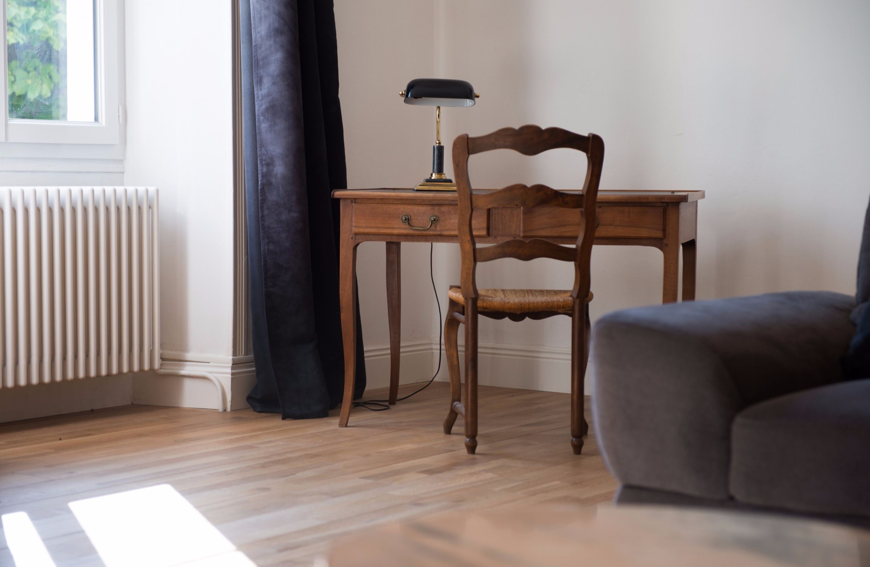 Uma mesa de madeira com uma lâmpada, uma cadeira e um radiador num quarto com soalho de madeira e uma janela com cortinas.
