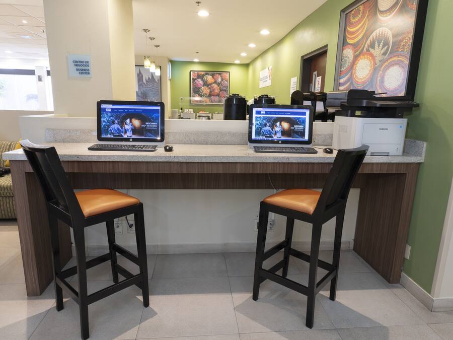 Uma área de recepção com dois bancos altos, um balcão com dois computadores HP, uma impressora Samsung e um grande painel decorativo.