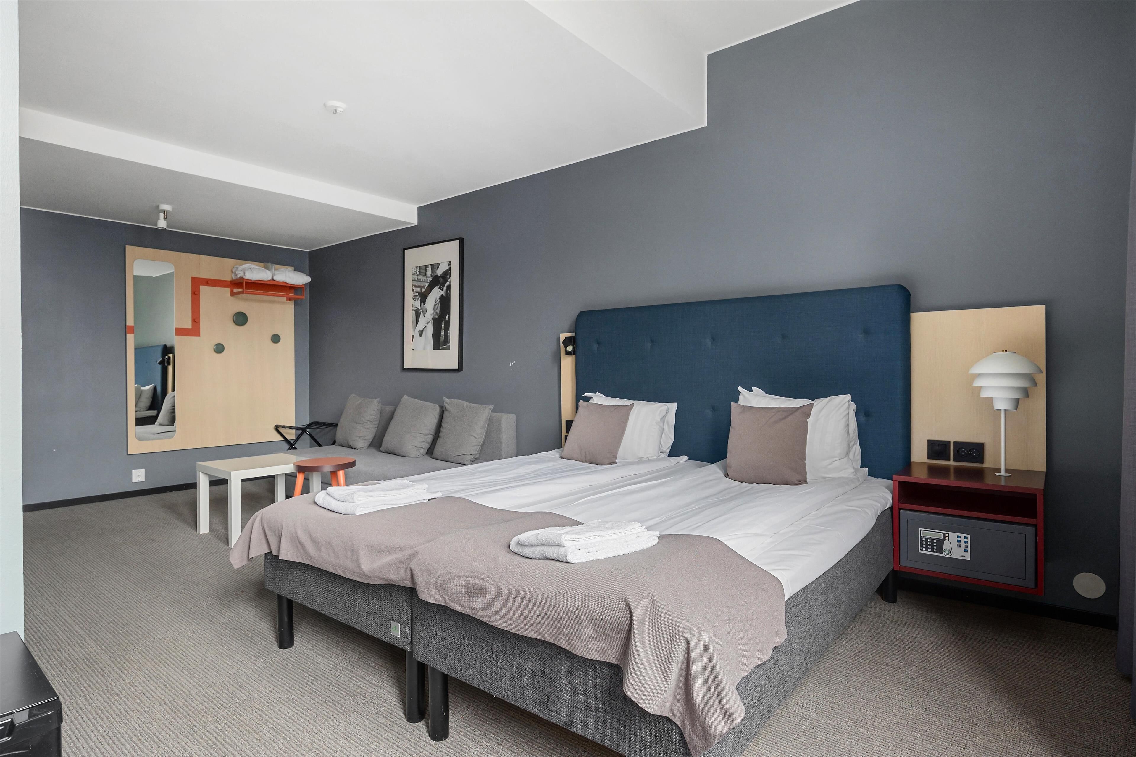 Quarto de hotel moderno com uma cama grande, uma mesa de cabeceira, um criado-mudo com abajur e um espelho na parede.