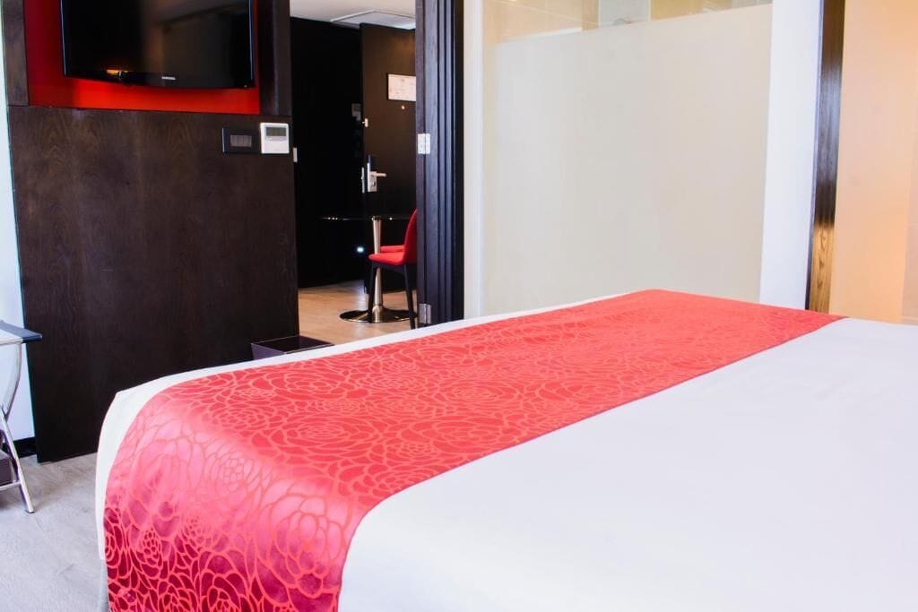 Quarto de hotel com uma cama grande, uma cadeira vermelha, uma televisão na parede e uma porta que dá para outro quarto.