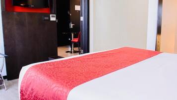 Quarto de hotel com uma cama grande, uma cadeira vermelha, uma televisão na parede e uma porta que dá para outro quarto.