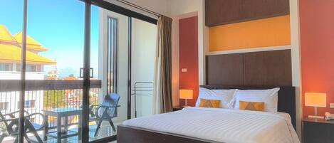Quarto de hotel com uma cama grande, duas luminárias de cabeceira, vista para um edifício com telhado amarelo e uma varanda com cadeiras.