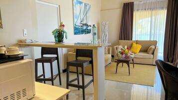 Quarto de hotel com cozinha compacta, área de refeições com cadeiras, sofá e uma janela grande com cortinas.