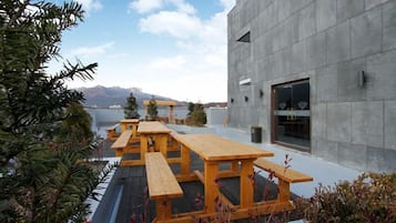 Uma área de estar externa com mesas de piquenique de madeira, um edifício moderno com uma grande janela e vista para montanhas ao fundo.