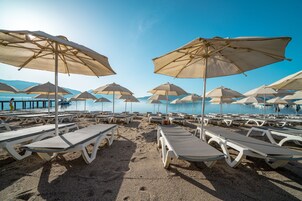 Uma praia com fileiras de espreguiçadeiras e guarda-sóis, piscina e montanhas ao fundo.