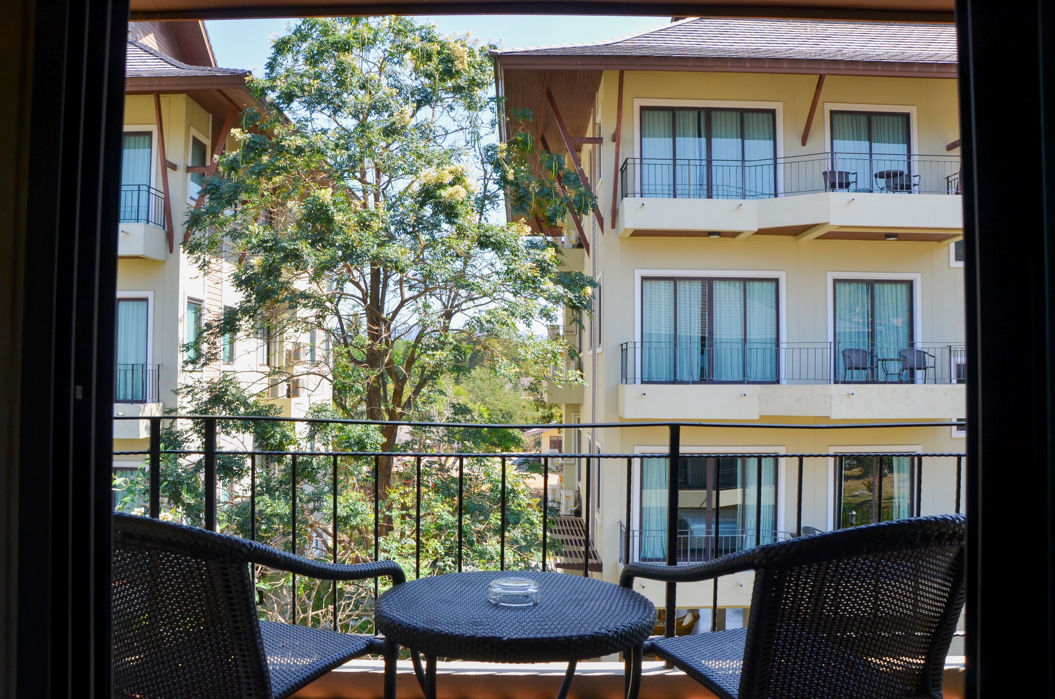 Uma varanda com móveis de vime, uma mesa de vidro e vista para um edifício com várias varandas e janelas.