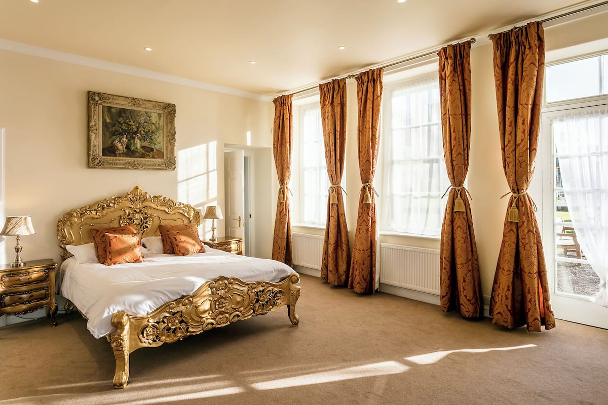Um quarto luxuoso com uma cama de dossel ricamente decorada, um quadro clássico e grandes janelas com cortinas.