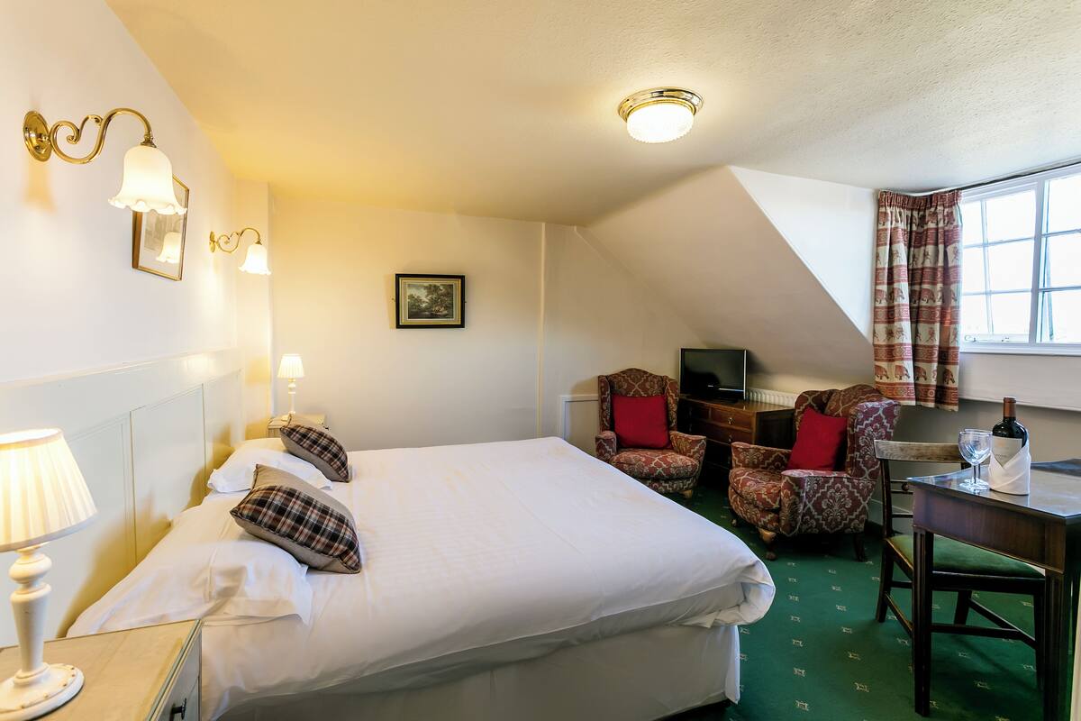 Quarto de hotel com cama grande, duas poltronas, escrivaninha, televisão e janela com cortinas.