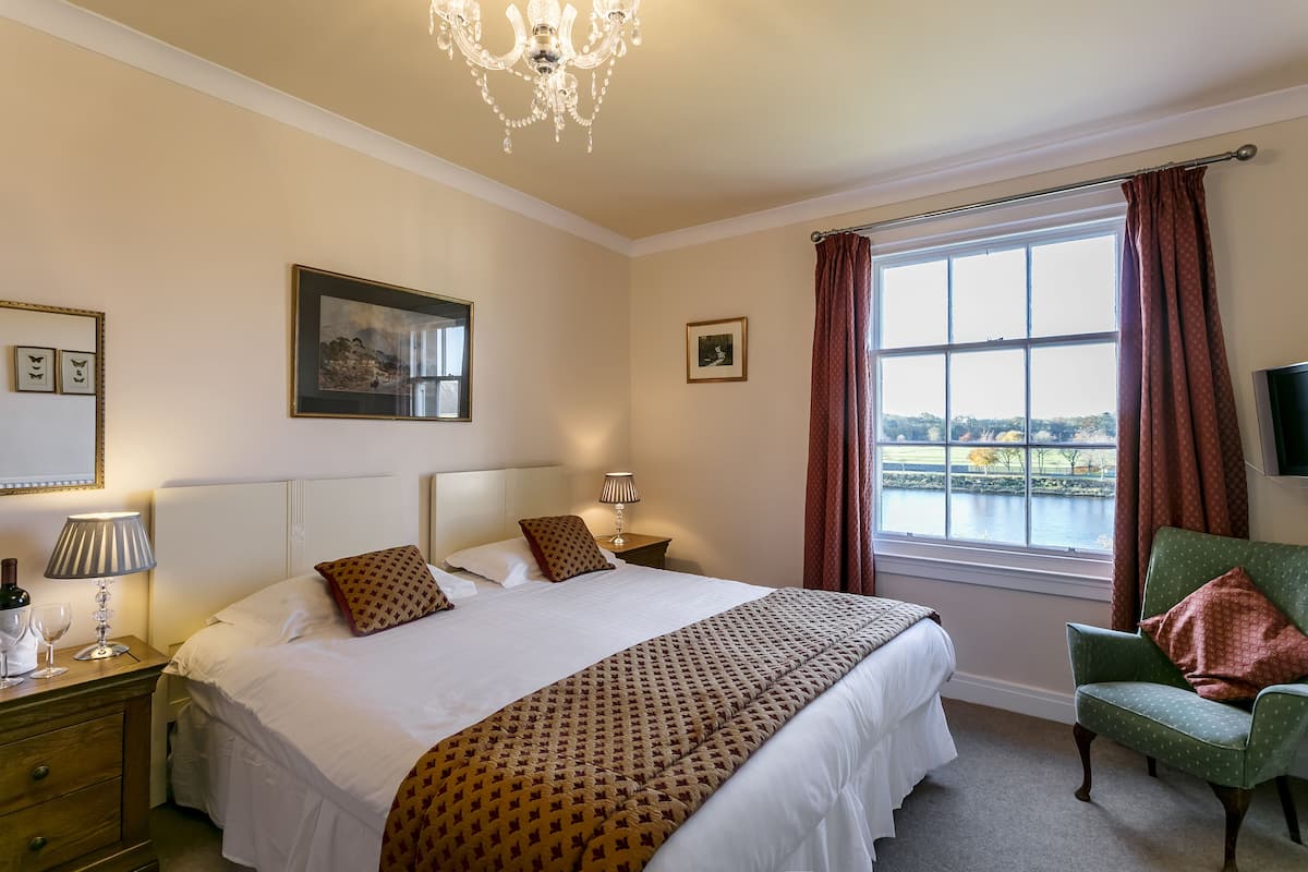 Quarto de hotel bem iluminado, com uma cama grande, uma cadeira, uma mesa de cabeceira com abajur e vista para a água através da janela.