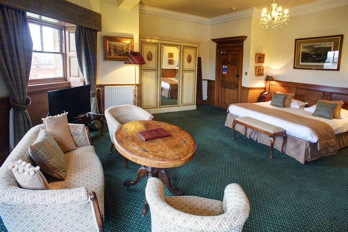 Quarto de hotel com uma cama, duas poltronas, uma mesa redonda de madeira, televisão e uma janela grande com cortinas.