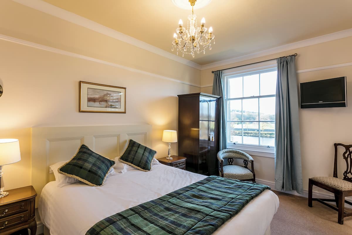 Quarto de hotel com uma cama grande, um lustre, uma televisão e uma janela com cortinas.