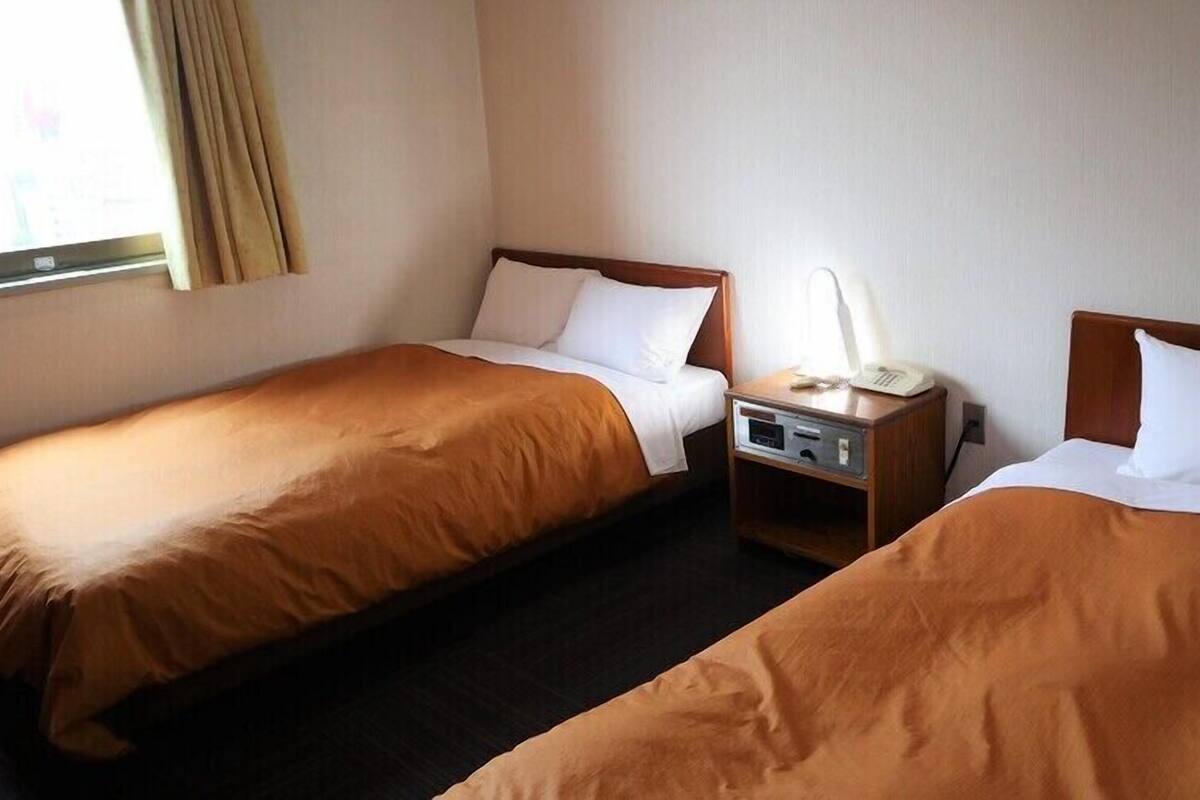 Quarto de hotel com duas camas de solteiro, uma mesa de cabeceira com abajur e uma janela com cortinas.