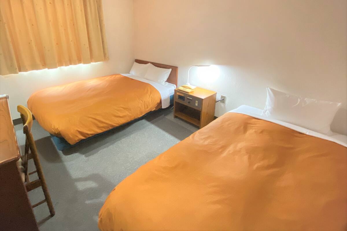 Quarto de hotel com duas camas, uma mesa de cabeceira, uma luminária e uma janela com cortinas.