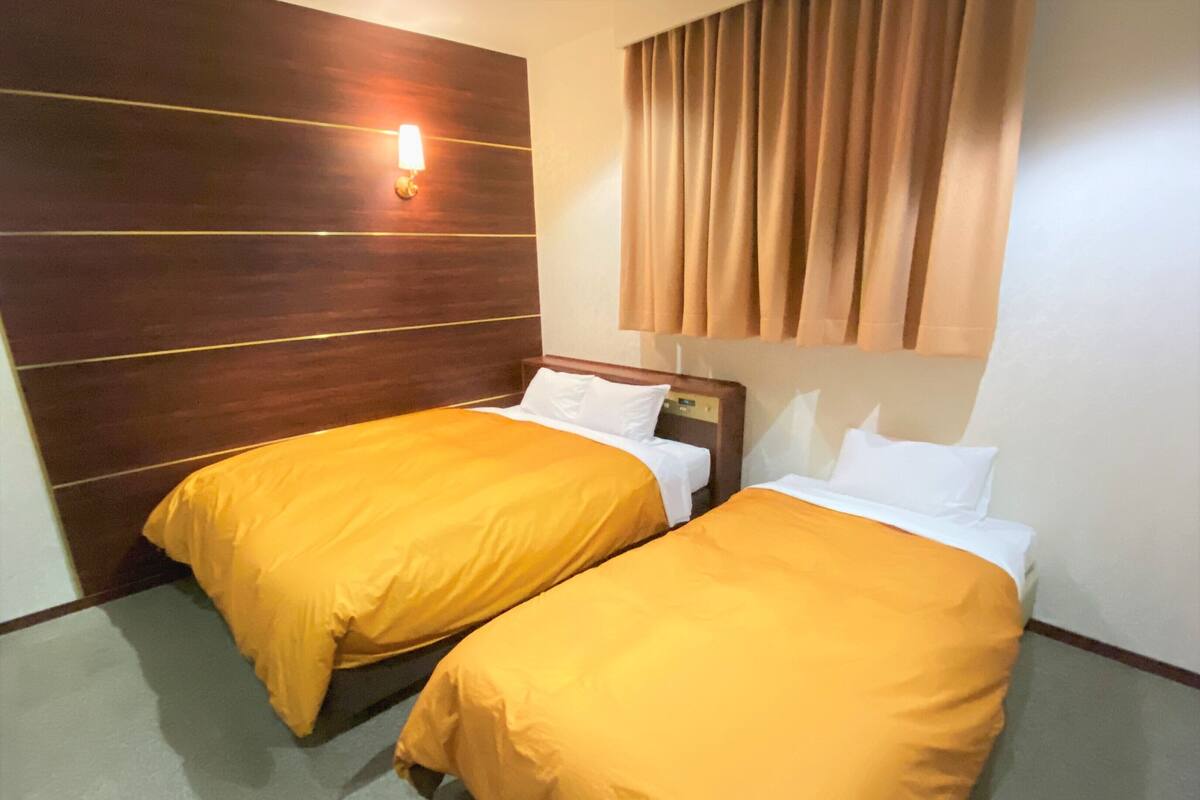 Quarto de hotel com duas camas de solteiro, cabeceiras de madeira, uma luminária fixada na parede e uma janela com cortinas.