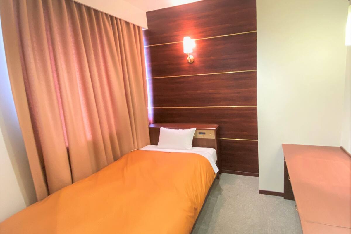 Quarto de hotel com cama, mesa de cabeceira, cabeceira de madeira, luminária na parede e janela com cortinas.