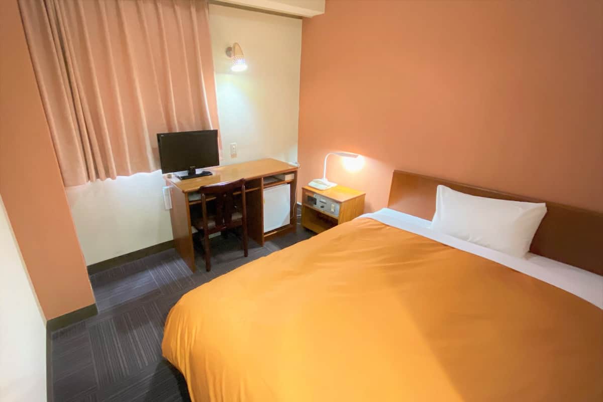 Quarto de hotel com cama, escrivaninha e cadeira, televisão, mesa de cabeceira com luminária e janela com cortinas.