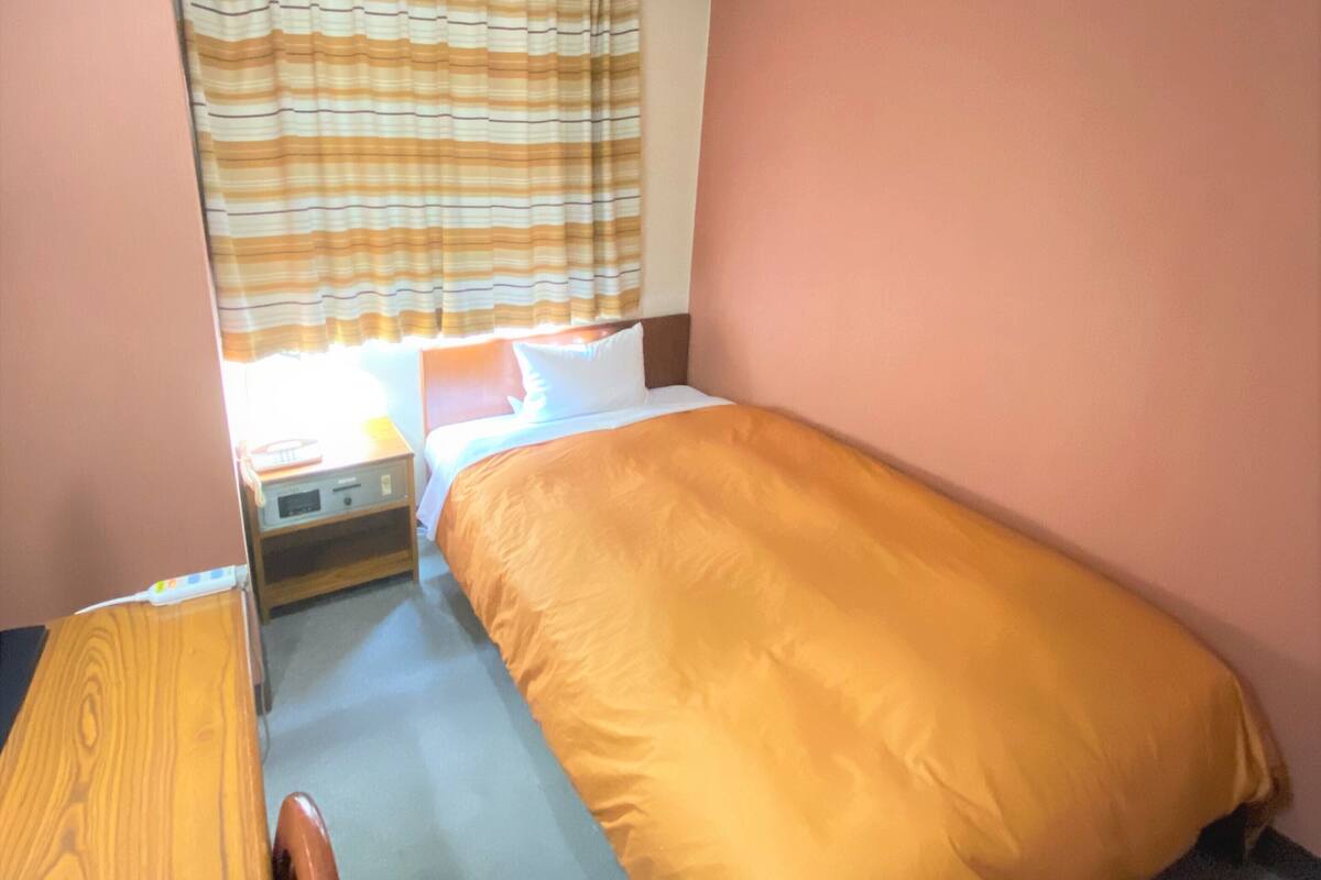 Quarto pequeno com uma cama de solteiro, mesa de cabeceira de madeira e cortina listrada.