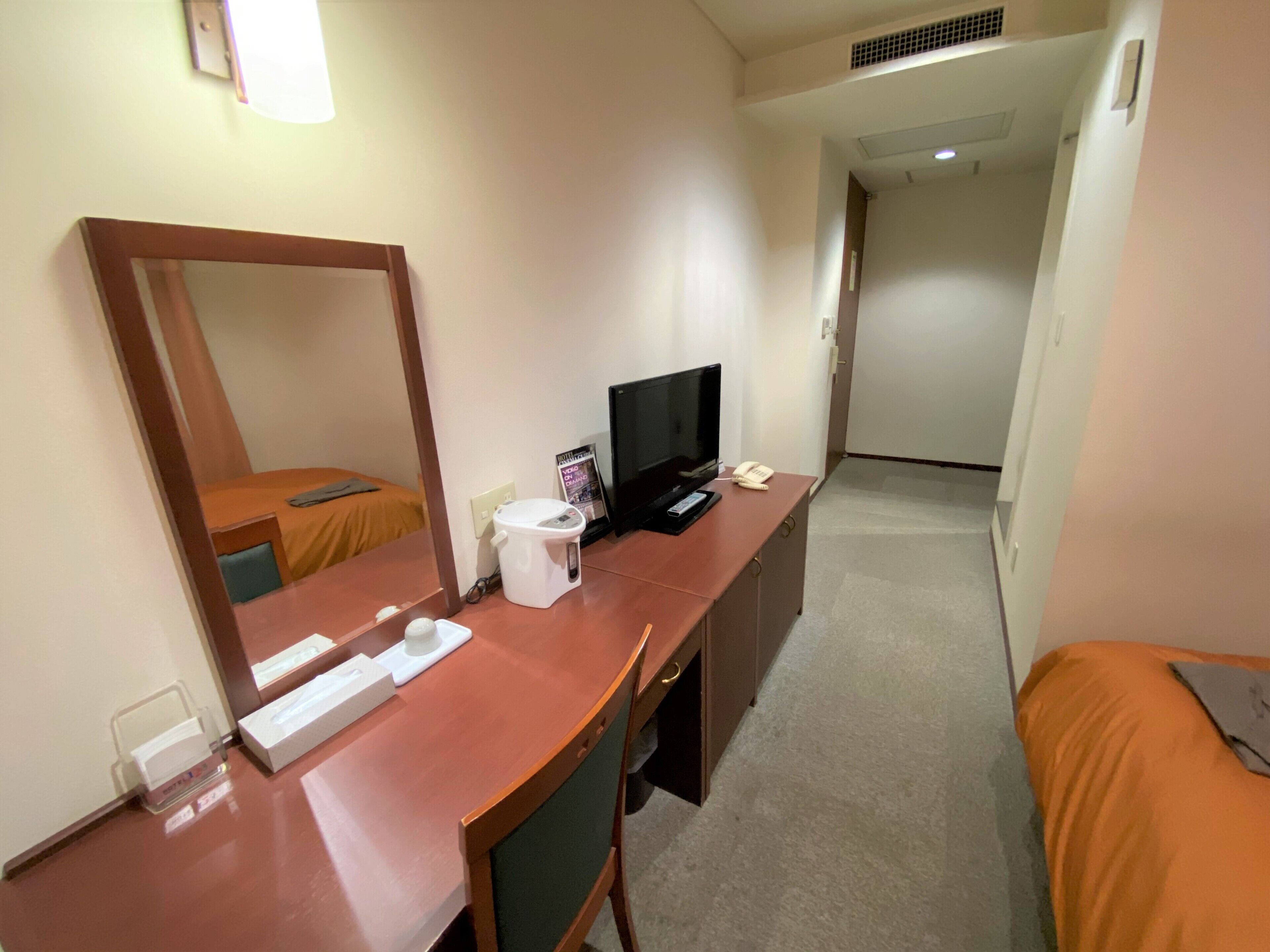 Quarto de hotel com cama, escrivaninha com espelho, televisão, telefone e uma pequena lixeira.