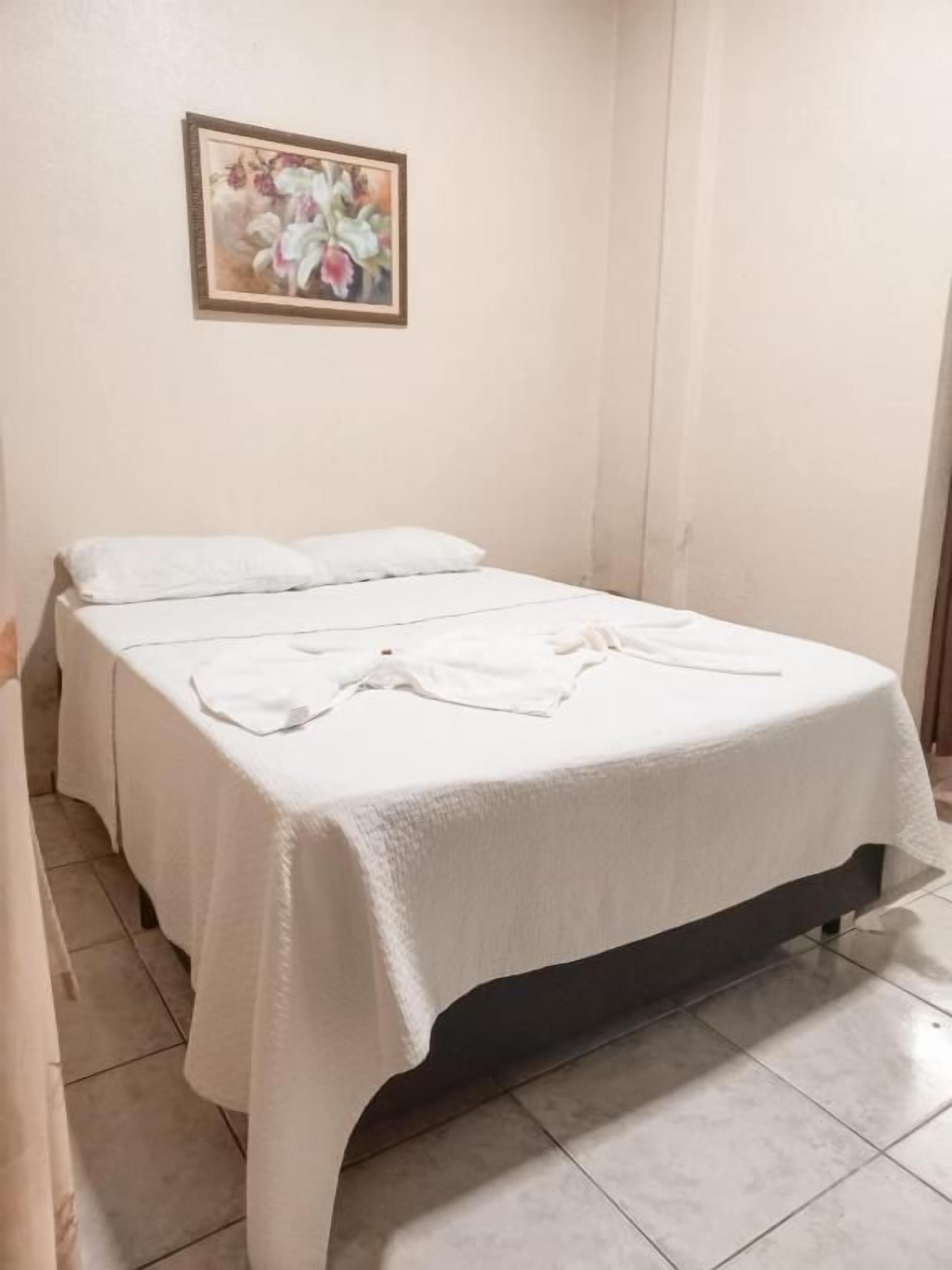 Uma cama de solteiro com colcha e travesseiros brancos, um quadro na parede e piso de azulejos.