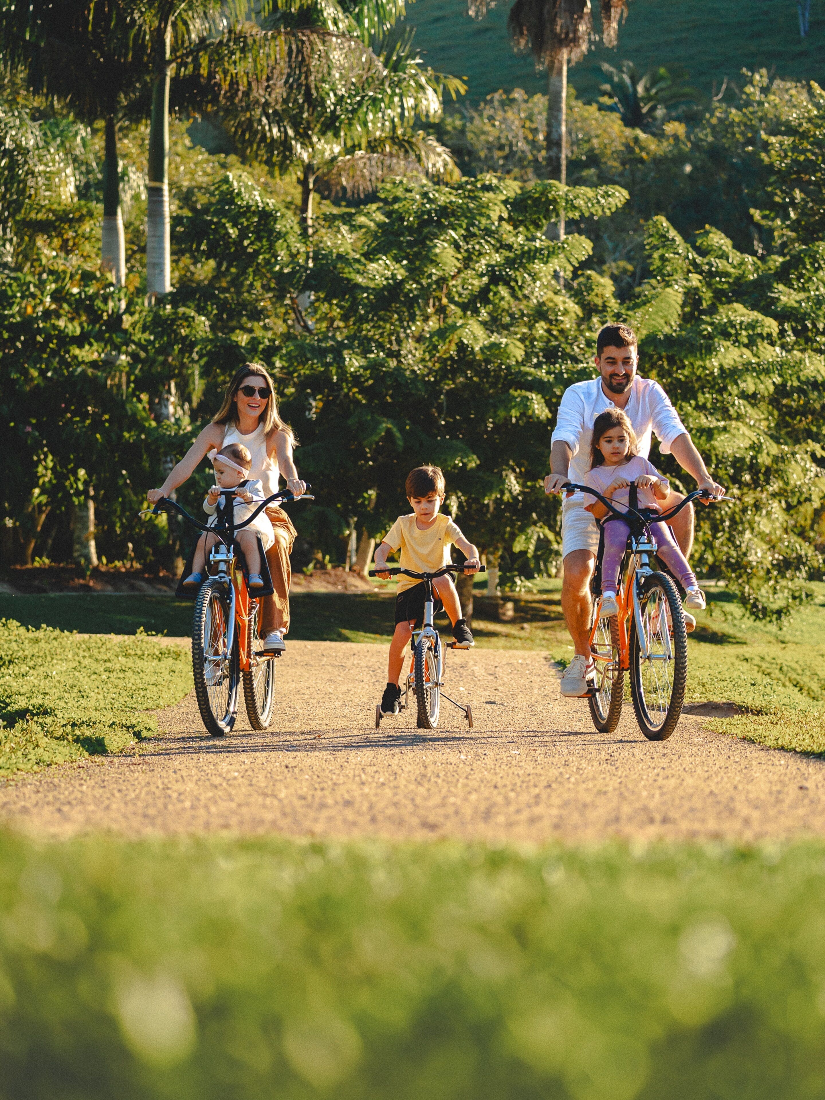 Uma família andando de bicicleta em um caminho de cascalho, cercada por vegetação exuberante e árvores.