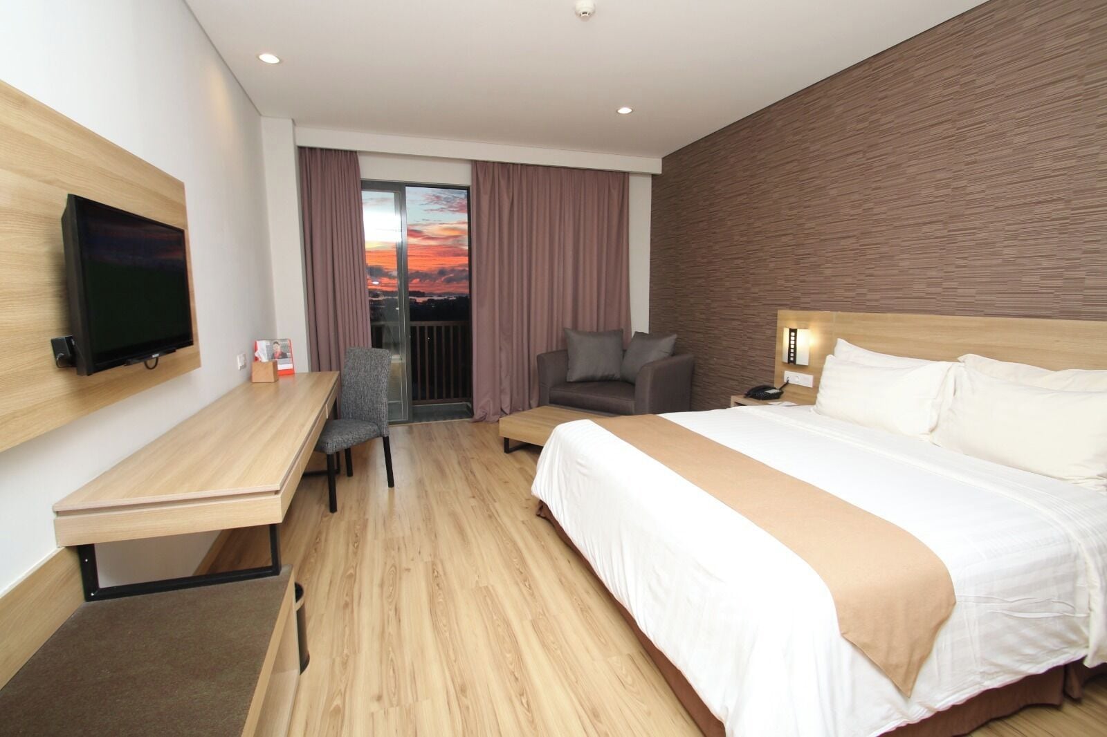 Quarto de hotel com cama, escrivaninha, cadeira e televisão fixada na parede.