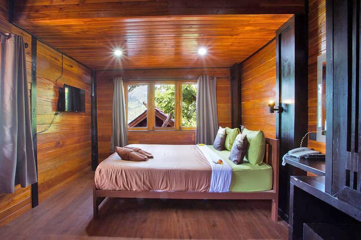 Quarto de cabana de madeira com cama, uma escrivaninha pequena e janela com cortinas.