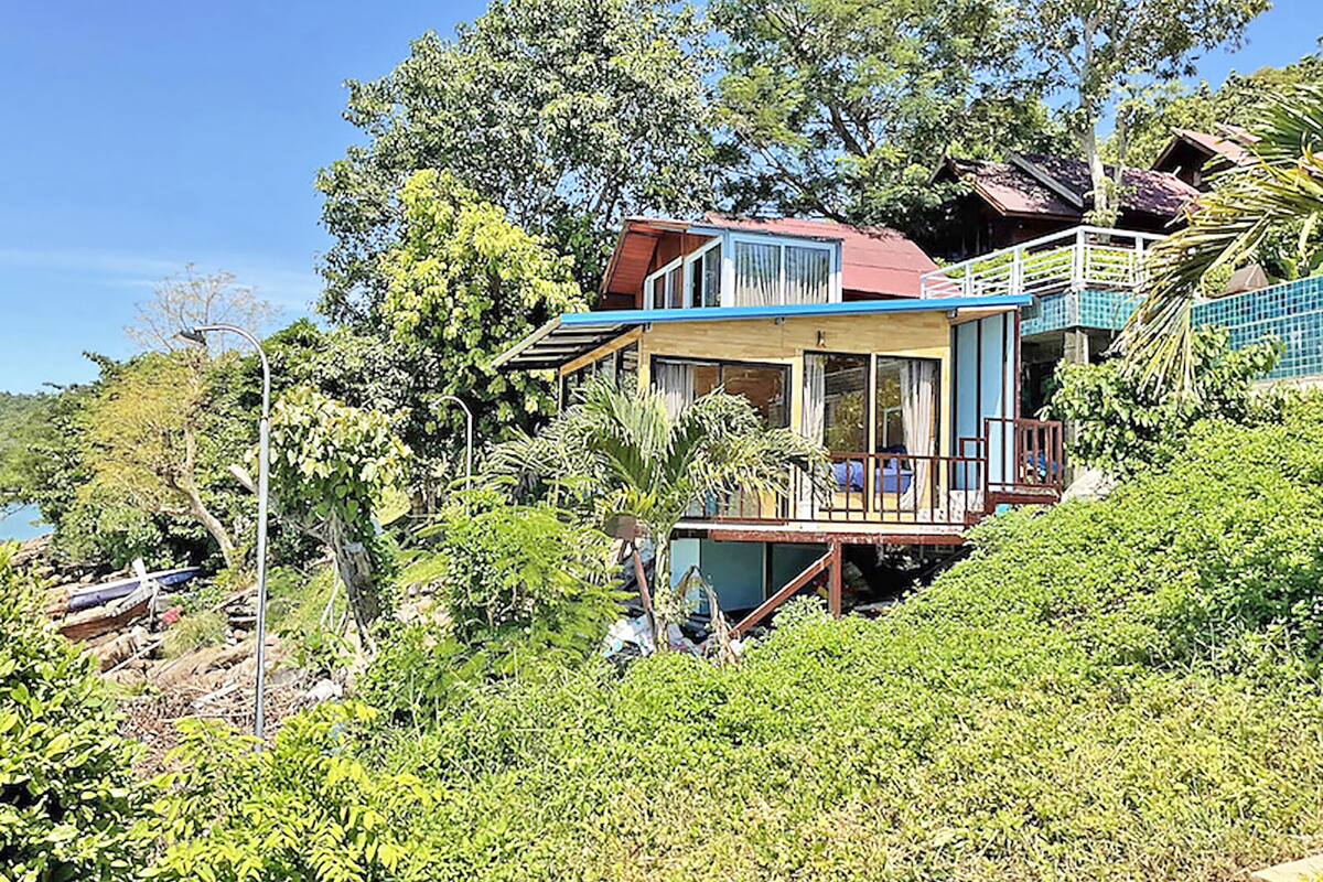 Uma casa de dois andares com uma varanda, rodeada por vegetação exuberante e árvores.
