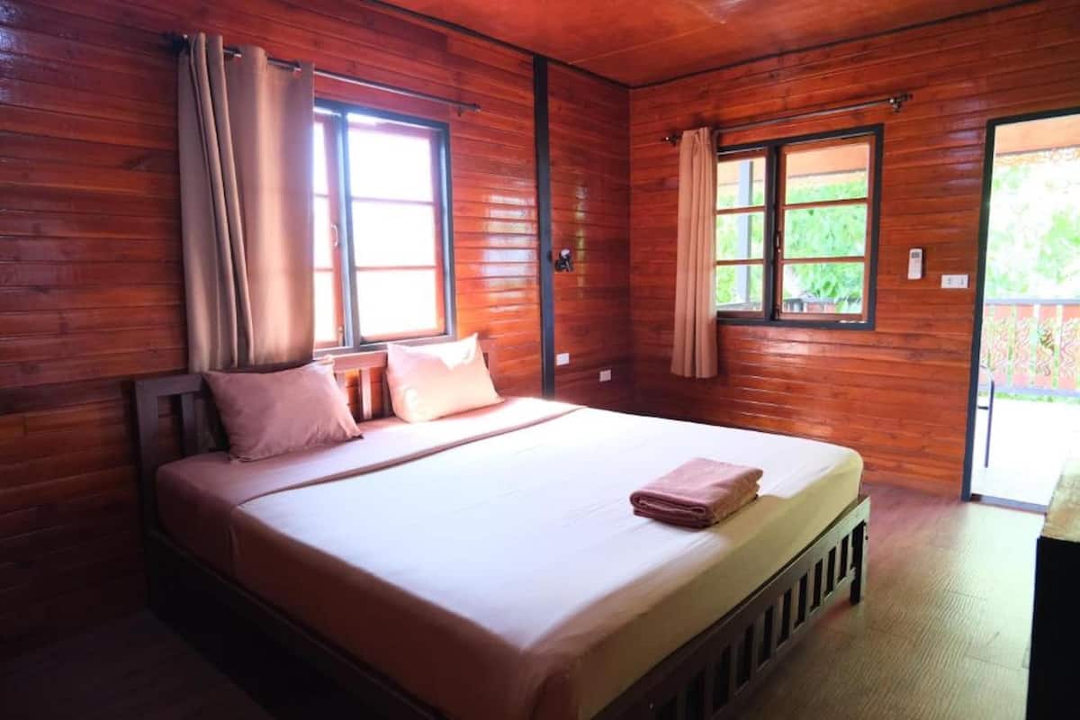 Quarto de cabana de madeira com cama, duas janelas com cortinas e uma porta que dá para o exterior.