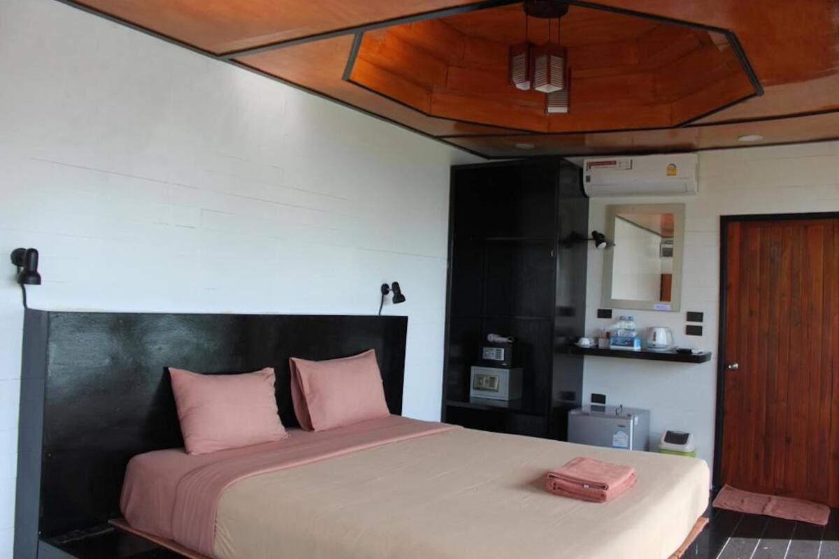 Quarto de hotel moderno com uma cama grande, teto de madeira, cabeceira preta, mesa de cabeceira, espelho e uma pequena escrivaninha com cadeira.